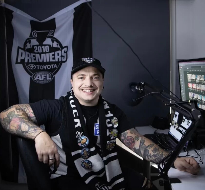 Luke Caruana sits at a table wearing collingwood hat and jumper, with a ...